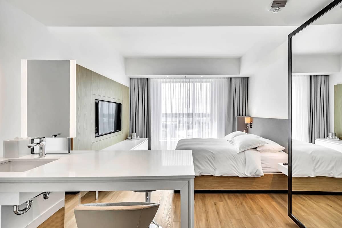 Quarto de hotel moderno com uma cama grande, uma escrivaninha com cadeira, televisão e banheiro com pia e espelho.