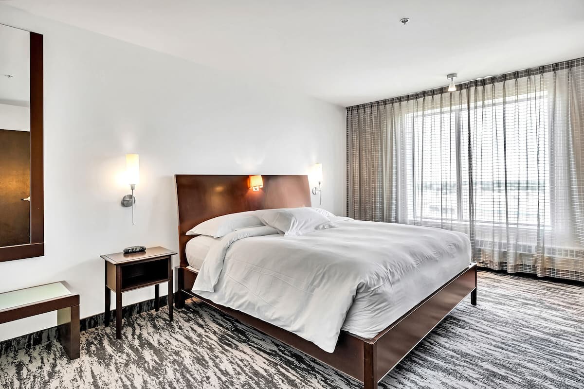 Quarto de hotel moderno com uma cama grande, mesas de cabeceira, um espelho e vista para o exterior através de cortinas leves.