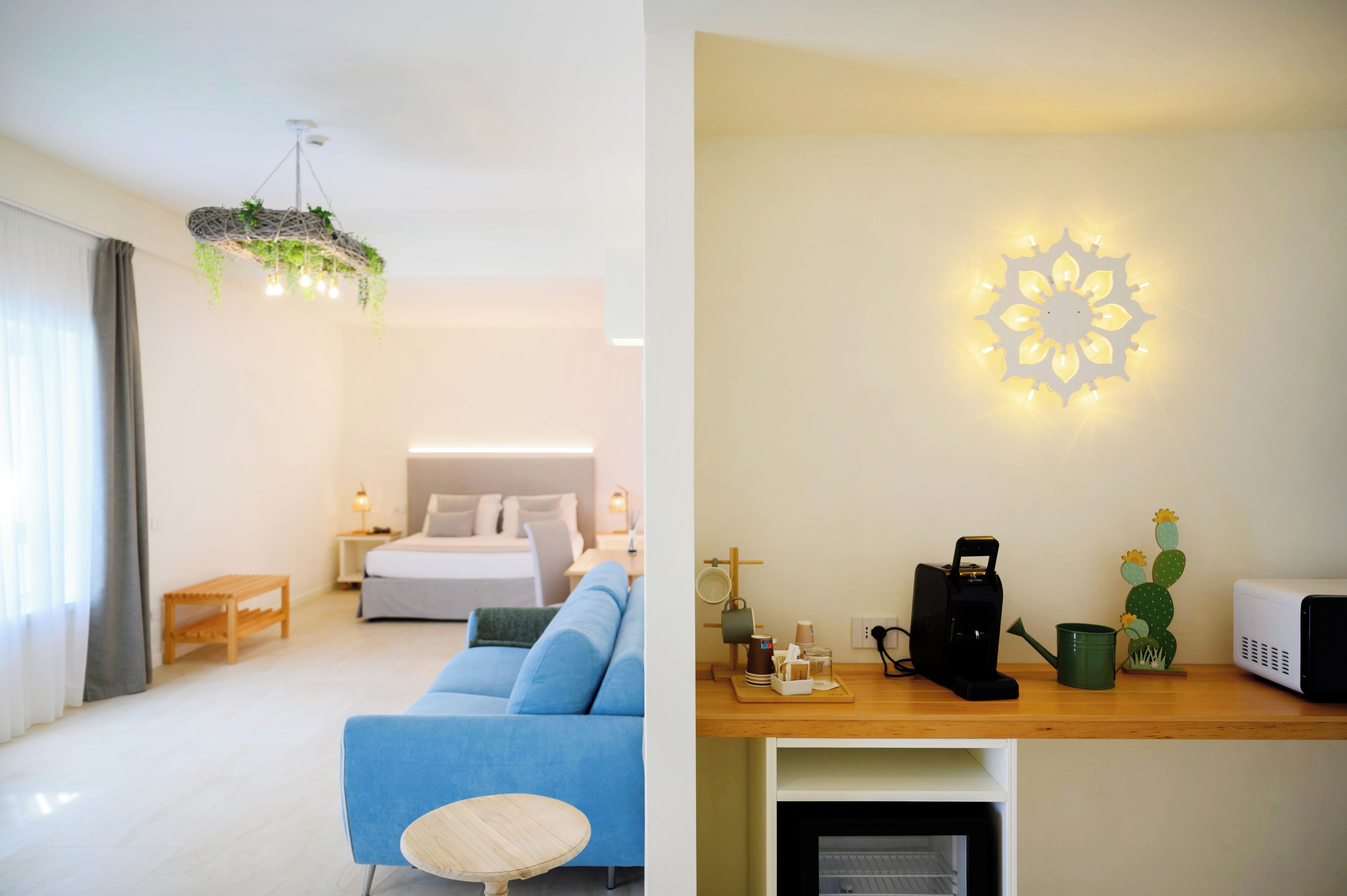 Quarto de hotel moderno com cama, sofá azul e área de refeições com cafeteira e decoração com cactos.