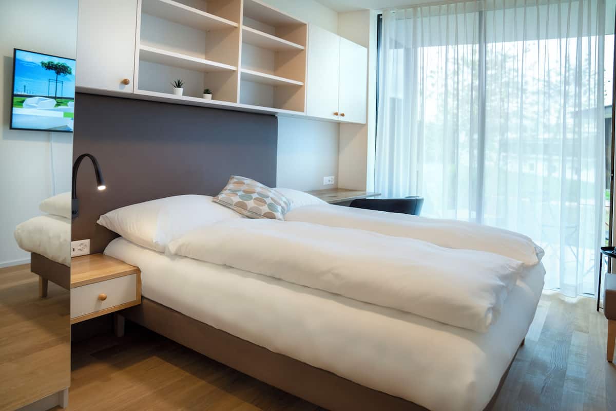 Um quarto moderno com cama, mesas de cabeceira, televisão e janela com cortinas leves.