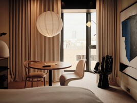 Quarto de hotel com uma cama, uma mesa redonda com cadeiras, uma janela grande com cortinas, um candeeiro pendente e uma escultura preta no chão.