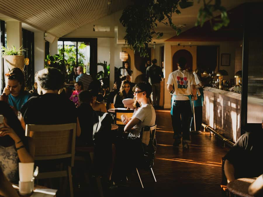 Um espaço amplo e movimentado para refeições no interior, com piso de madeira, plantas em vasos e um balcão de bar.