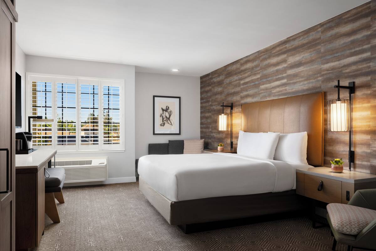 Quarto de hotel com uma cama grande, uma mesa de trabalho e uma cadeira. Há uma janela com persianas, um quadro emoldurado na parede e uma luminária fixada na parede.