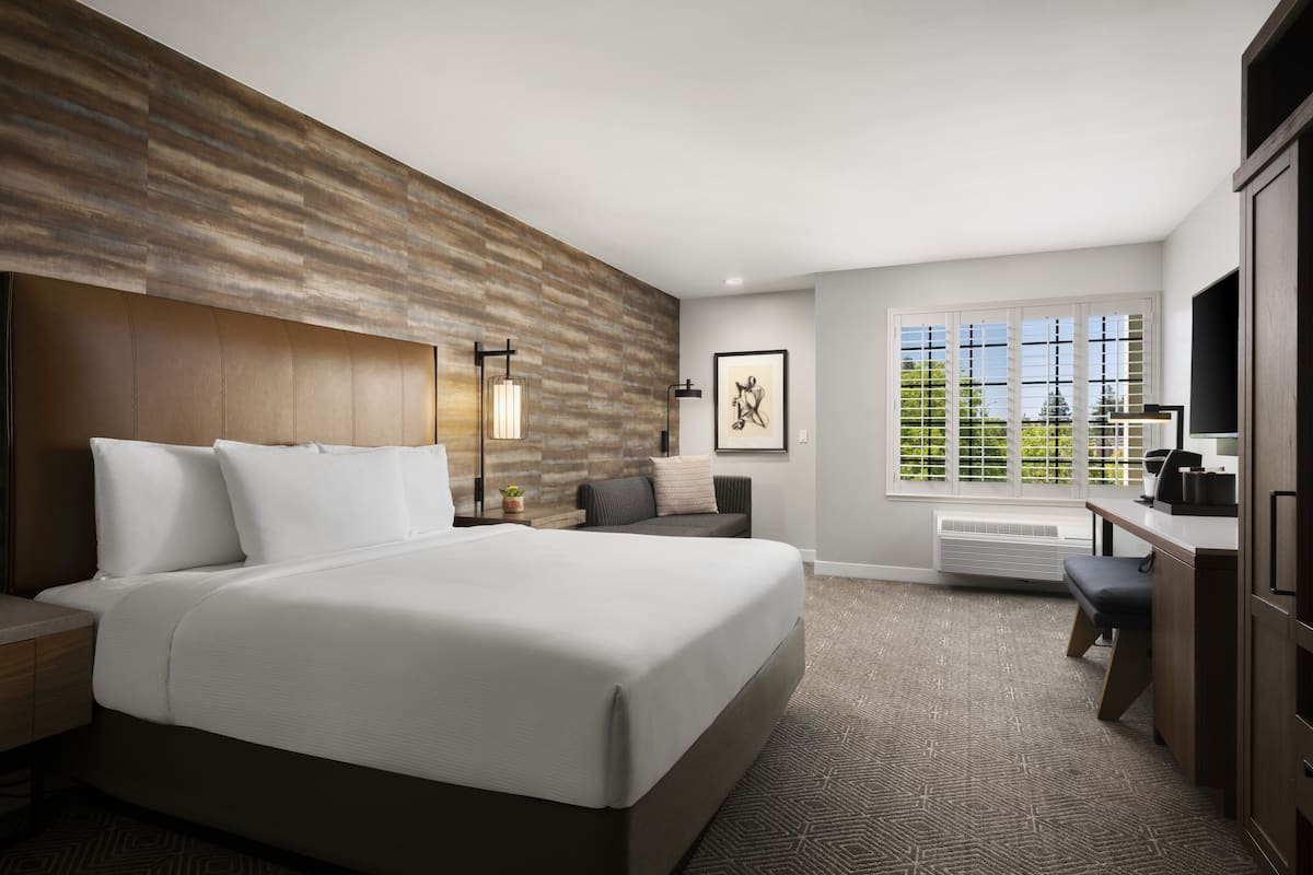 Quarto de hotel moderno com uma cama grande, uma escrivaninha e uma janela com vista para vegetação.