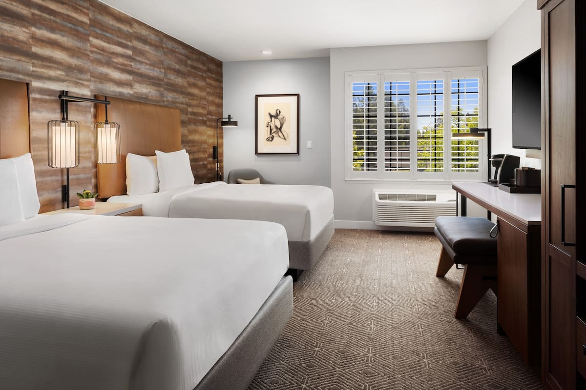 Quarto de hotel com duas camas, uma escrivaninha, televisão e janela com persianas.