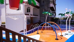 Uma área de recreação aquática com vários jatos de água e uma piscina, cercada por um edifício branco com varandas e palmeiras.