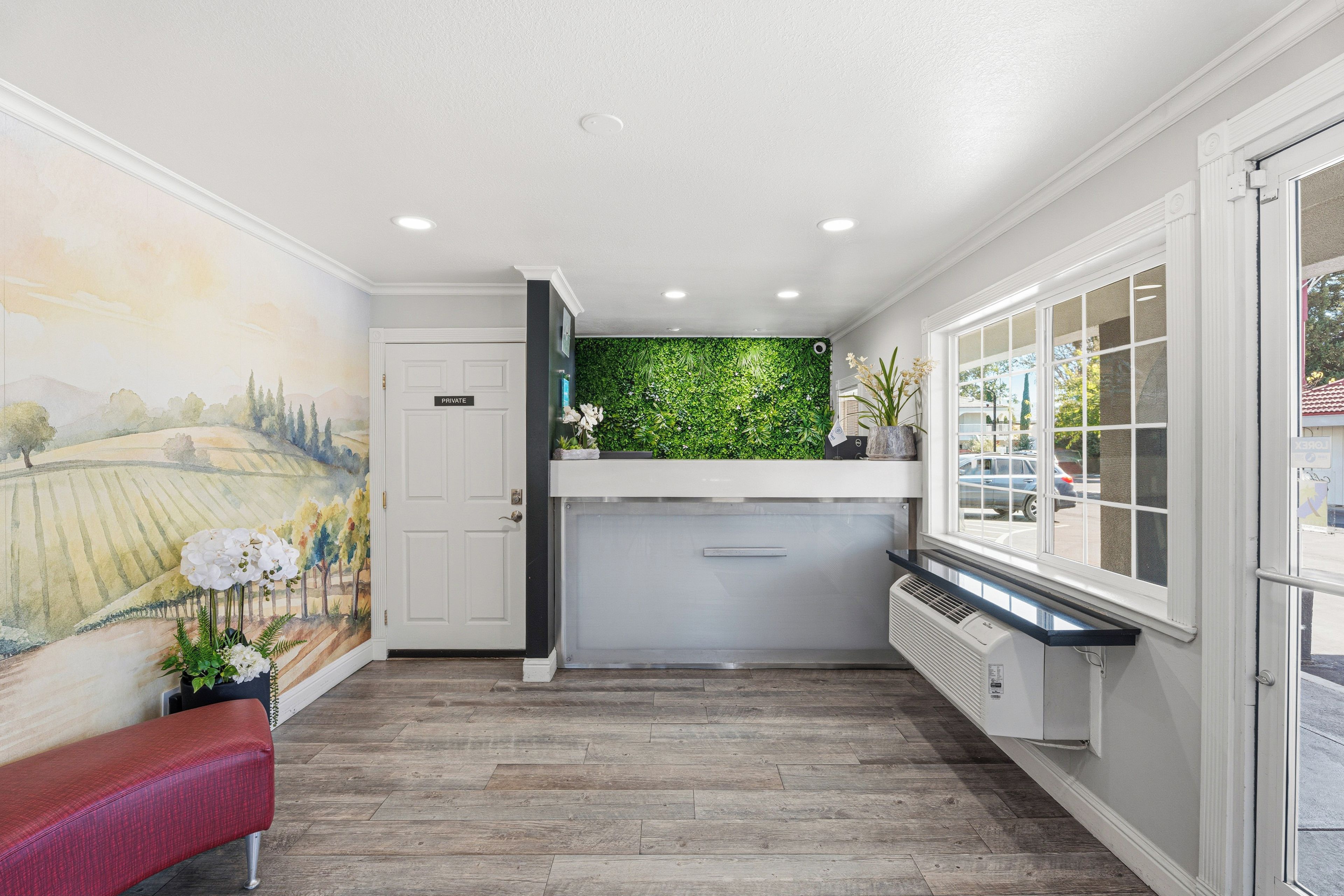 Quarto com piso de madeira, um banco, uma porta com a inscrição 'PRIVATE' e uma parede verde, além de uma janela com vista para o exterior.