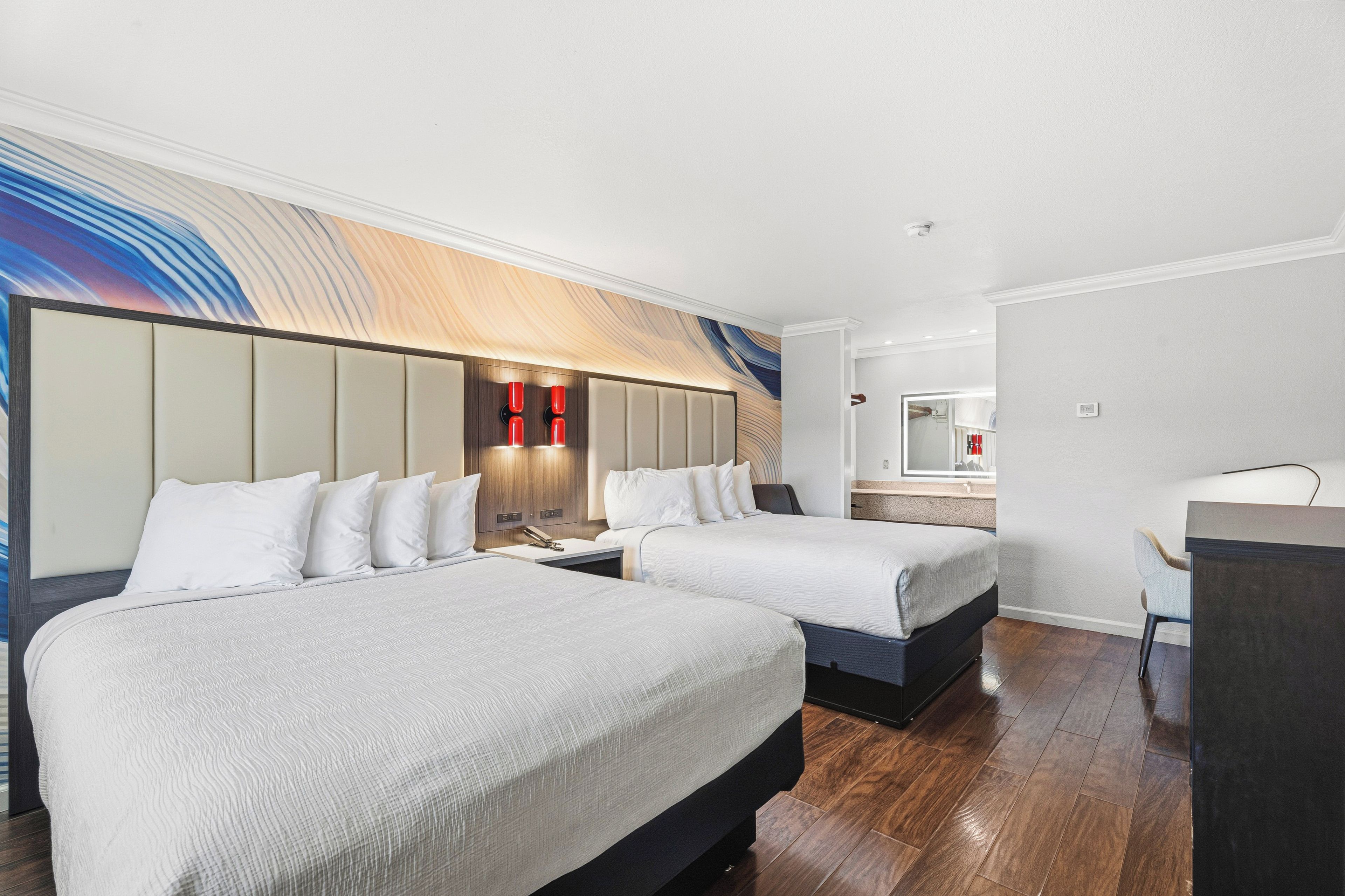Quarto de hotel com duas camas, uma mesa de trabalho e um banheiro visível ao fundo.