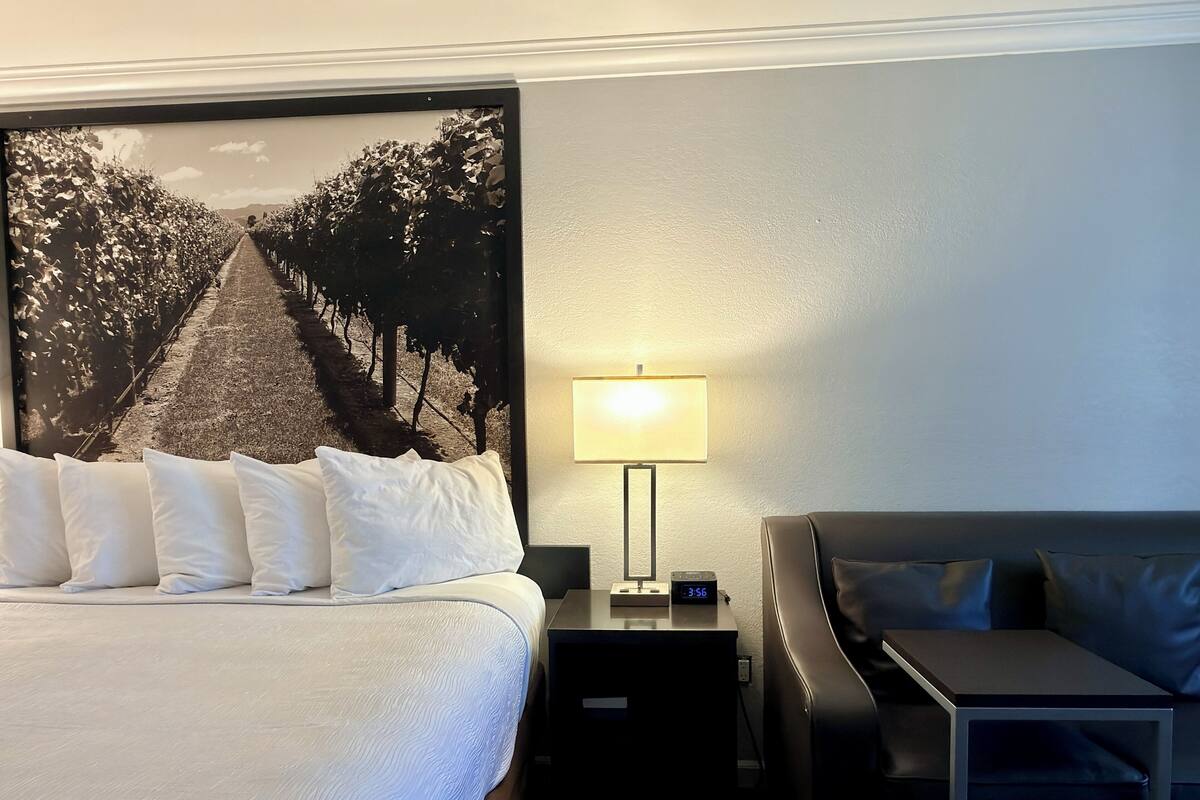 Quarto de hotel com uma cama grande, uma mesinha de cabeceira com abajur, um relógio digital, um sofá e um quadro pendurado na parede.