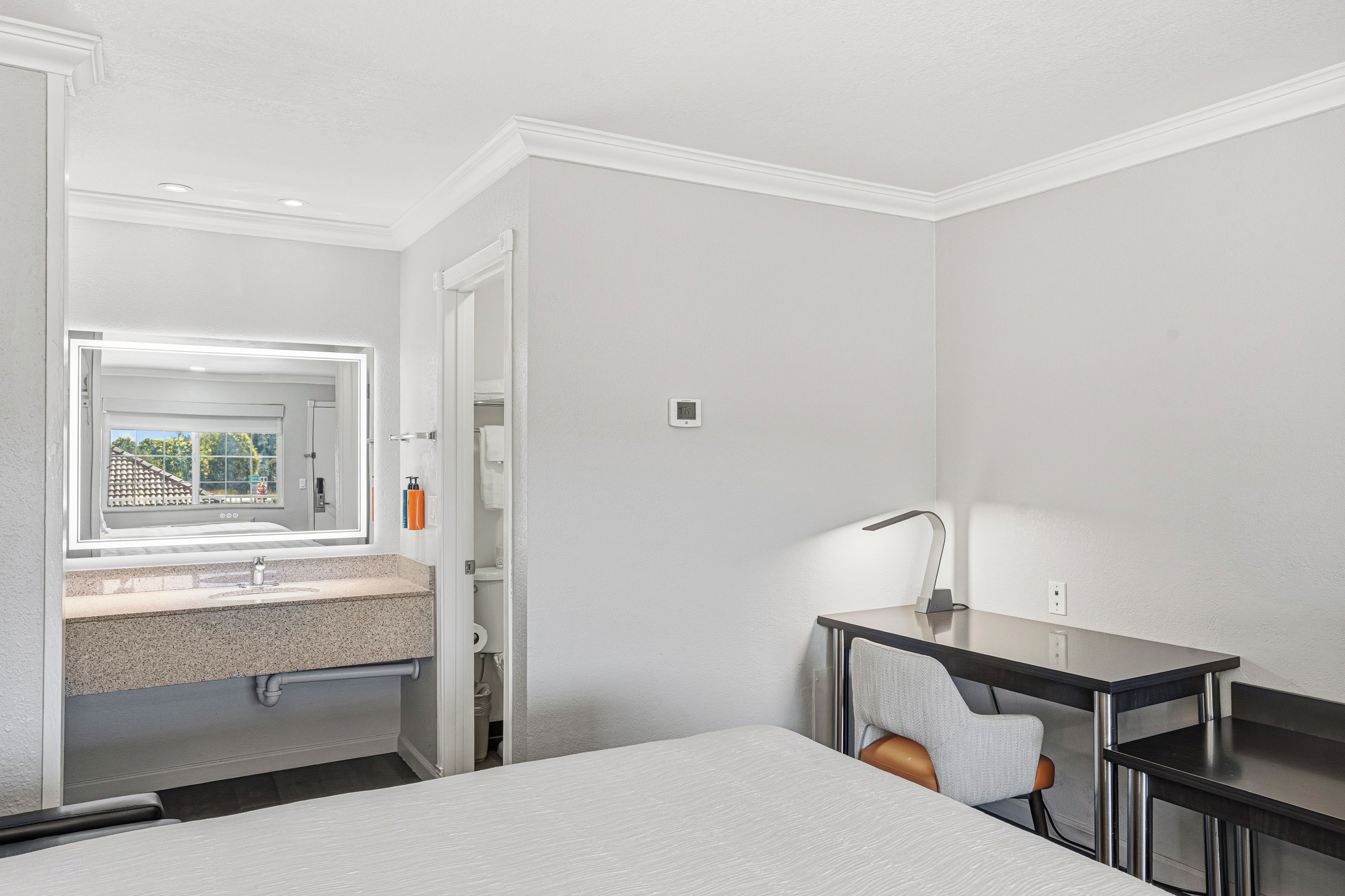 Quarto de hotel com cama, escrivaninha, cadeira, banheiro com pia e espelho, e vista para o exterior.