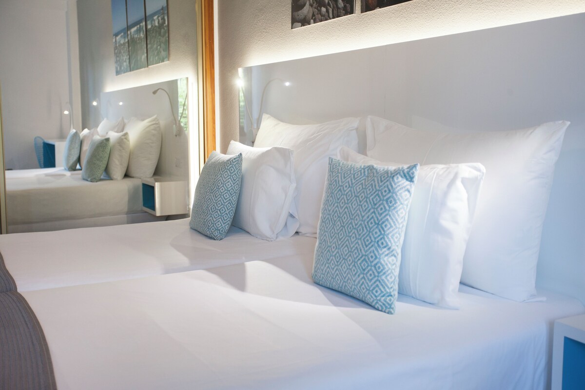 Quarto de hotel moderno com uma cama grande, várias almofadas e uma mesa de cabeceira.