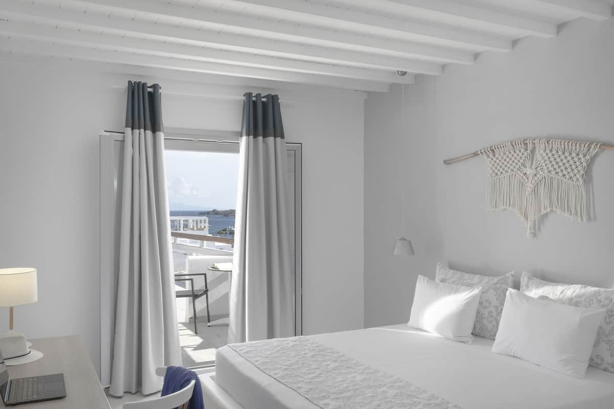 Quarto de hotel com cama, escrivaninha com laptop, cadeira e vista para o mar.
