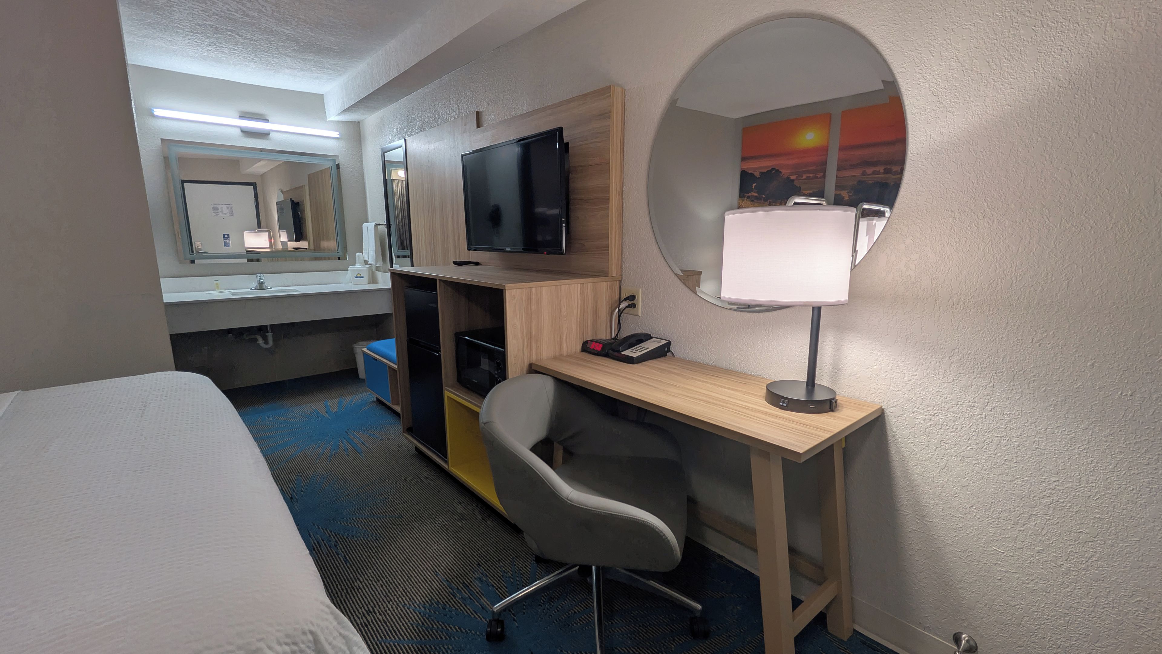 Quarto de hotel com cama, escrivaninha, cadeira, televisão e uma pintura do pôr do sol.