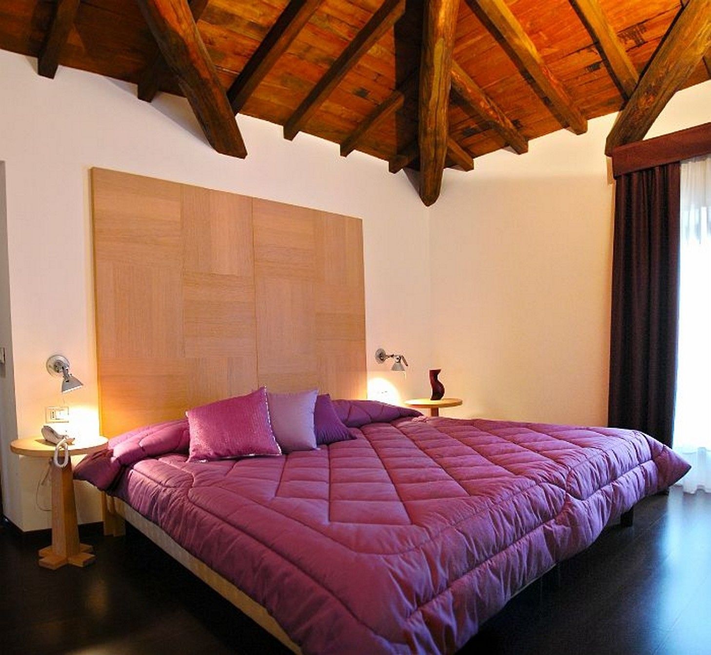Quarto com cama grande, cabeceira de madeira, duas mesas de cabeceira com luminárias e janela com cortinas.