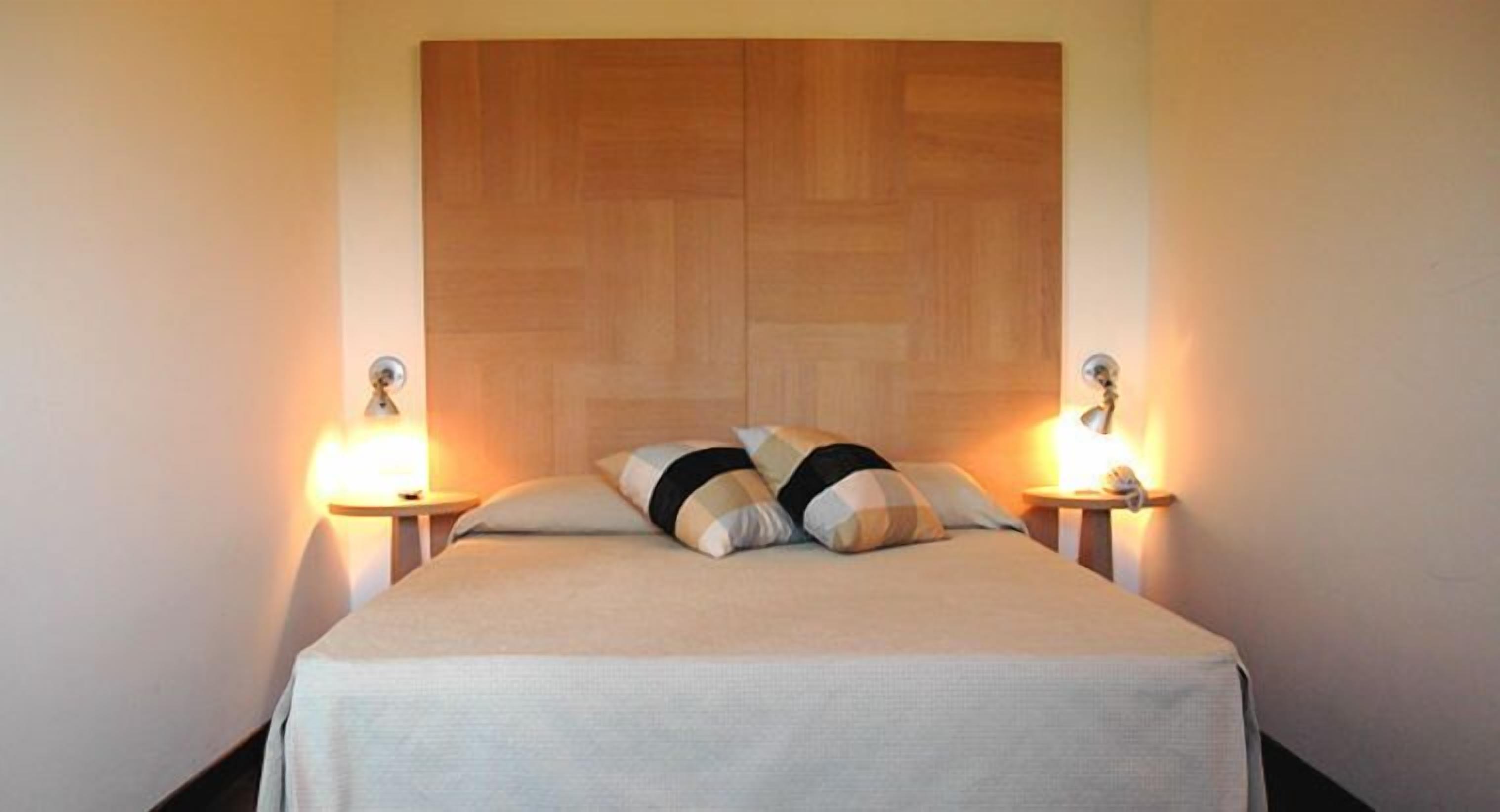 Cama bem arrumada com cabeceira de madeira, duas luminárias de apoio e um espalha-cama em tons neutros.