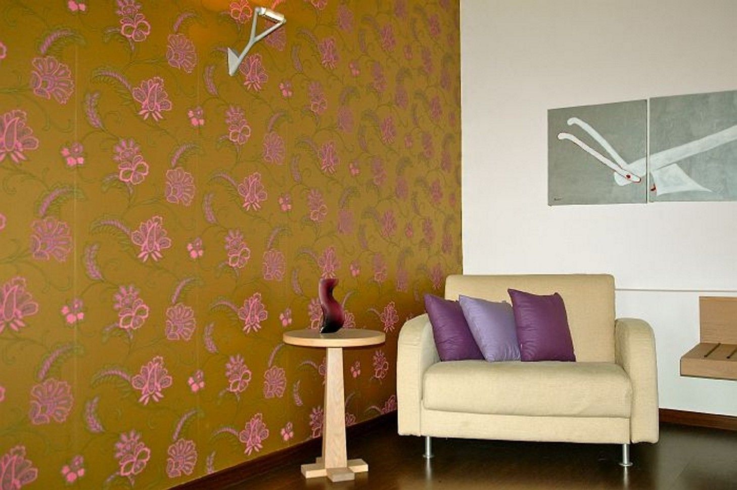 Um quarto com papel de parede floral, um sofá bege com almofadas roxas, uma pequena mesa lateral de madeira e uma pintura abstrata moderna na parede.