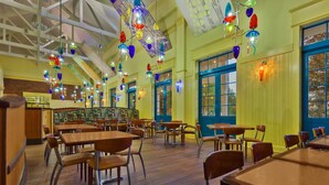 Um restaurante com mesas e cadeiras de madeira, decorações penduradas e coloridas, e janelas amplas.