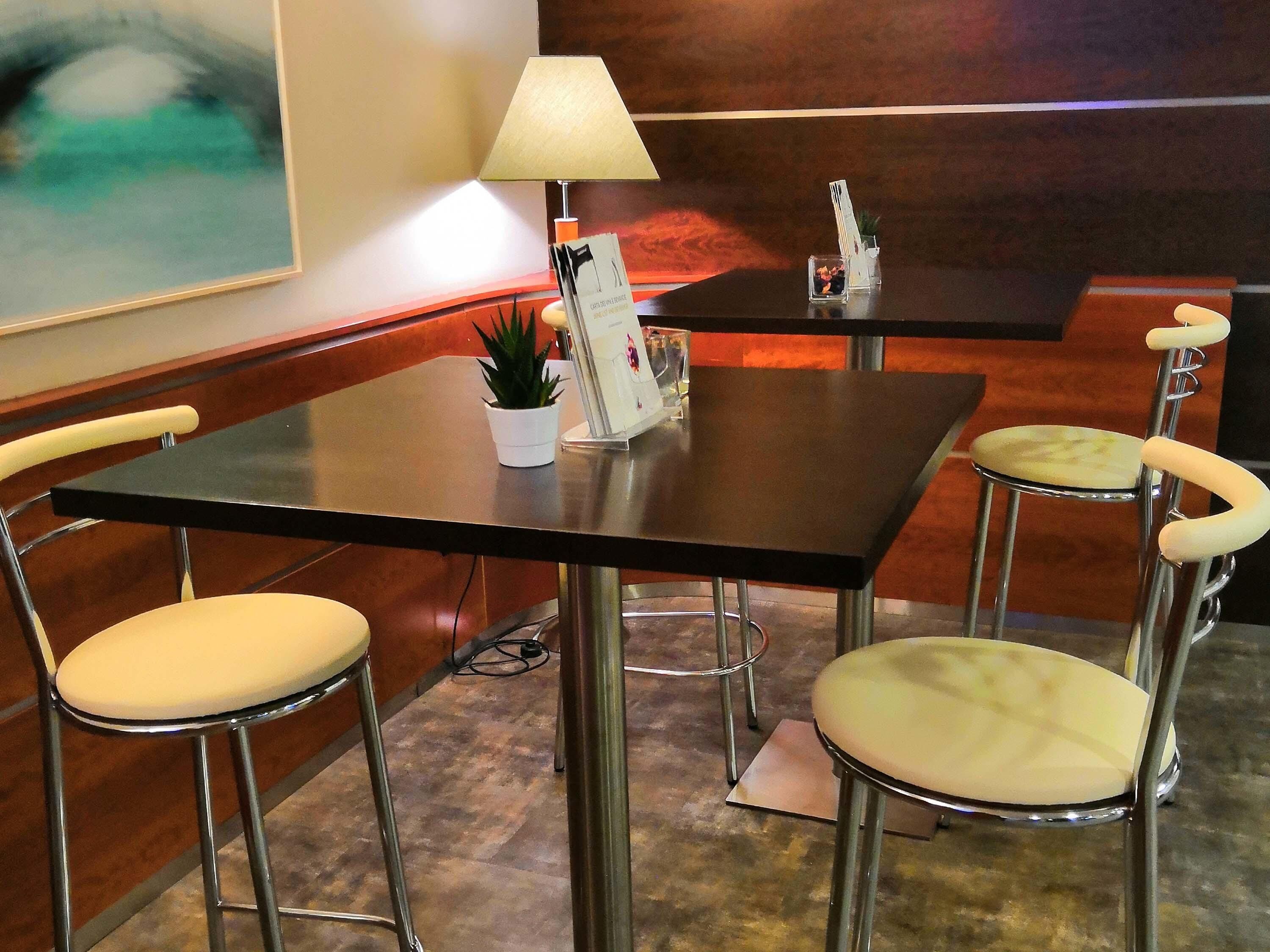 Uma área de recepção de hotel com balcão, dois bancos altos, um vaso de planta, uma luminária e um quadro na parede.