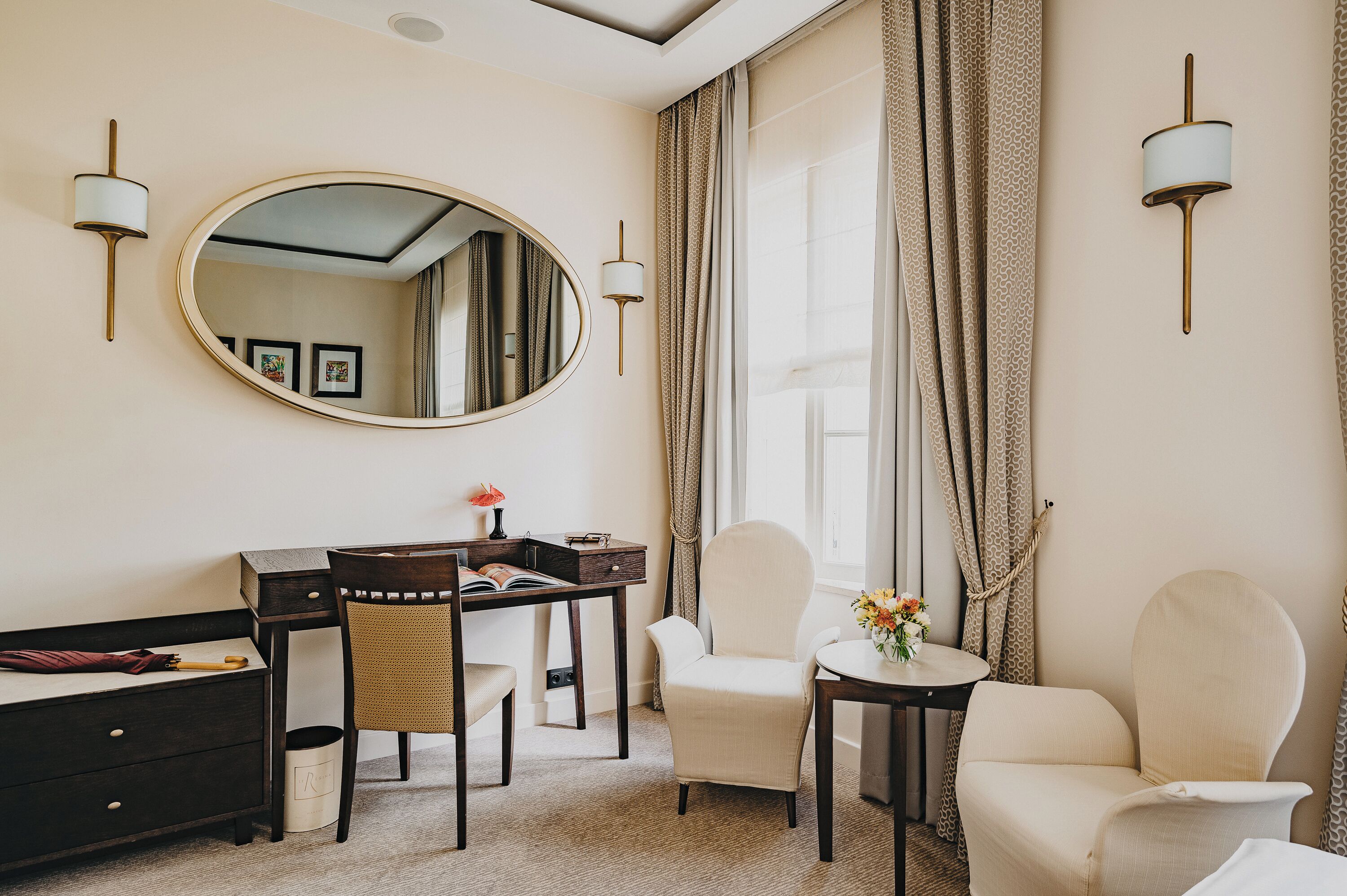 Quarto de hotel com escrivaninha, duas cadeiras, um espelho redondo e uma janela com cortinas.