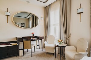Quarto de hotel com escrivaninha, duas cadeiras, um espelho redondo e uma janela com cortinas.