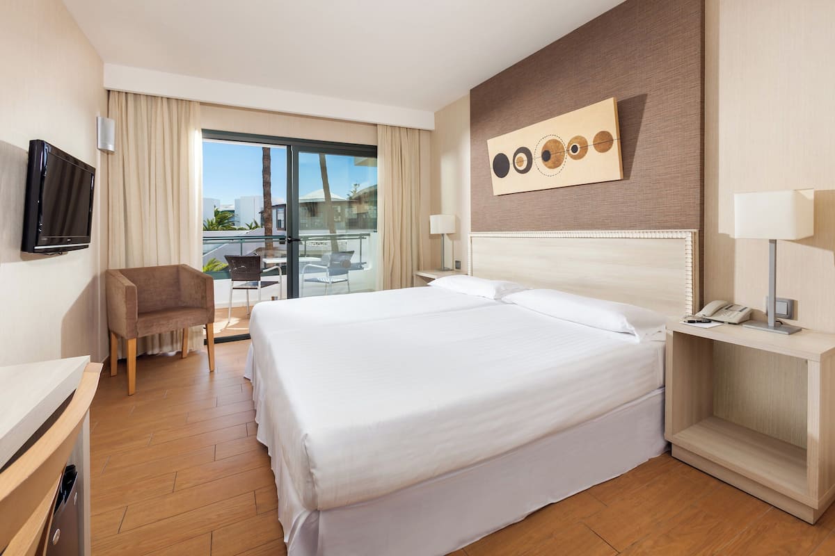Quarto de hotel com uma cama grande, televisão, escrivaninha com cadeira e varanda com vista.