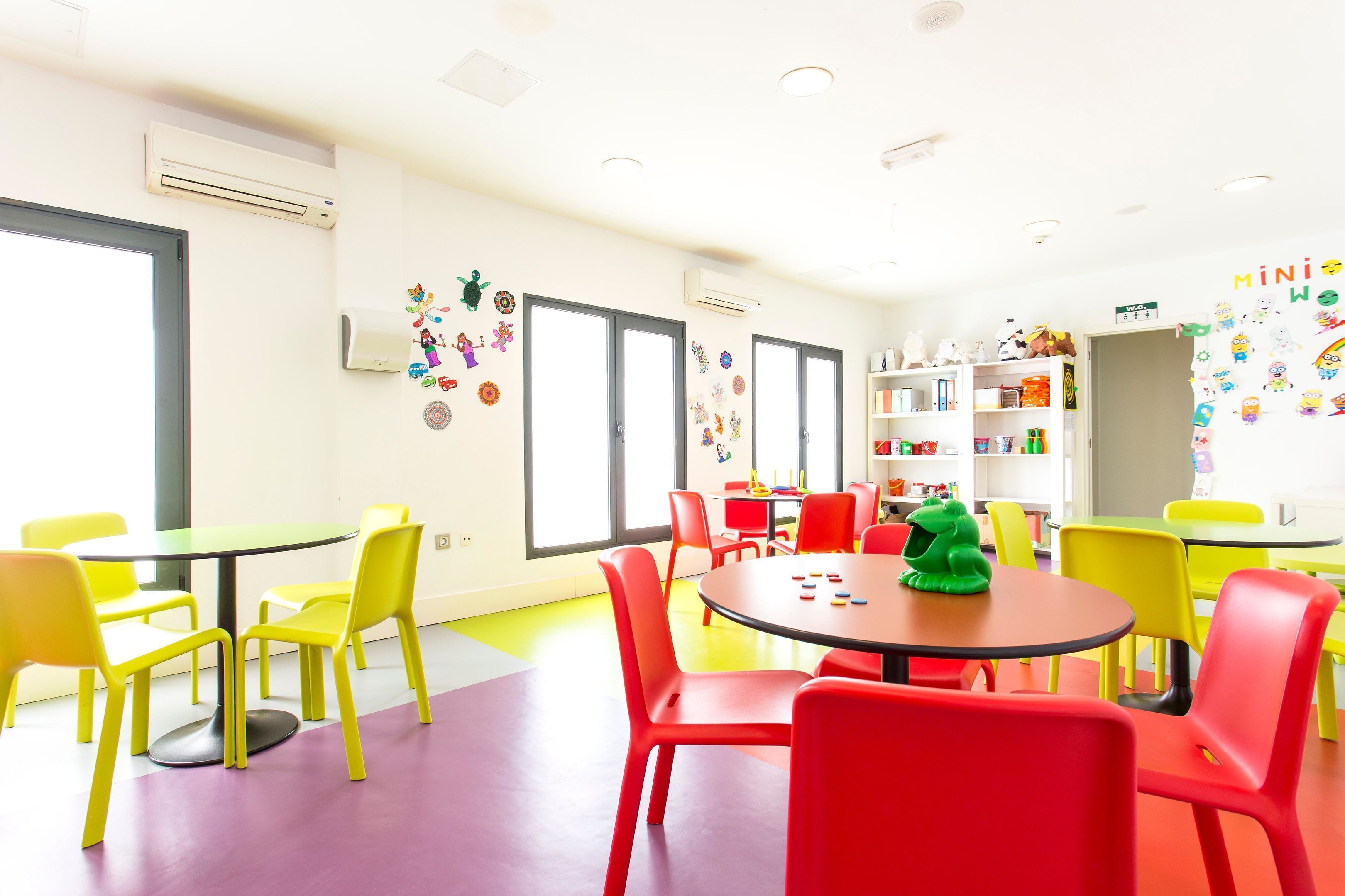 Uma sala de brinquedos infantil vibrante, com cadeiras amarelas e vermelhas, um brinquedo verde sobre uma mesa e decorações educativas nas paredes.