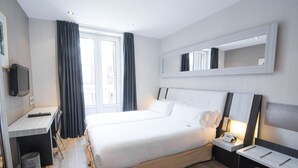 Quarto de hotel com cama, mesa de cabeceira, luminária, televisão e escrivaninha com cadeira.