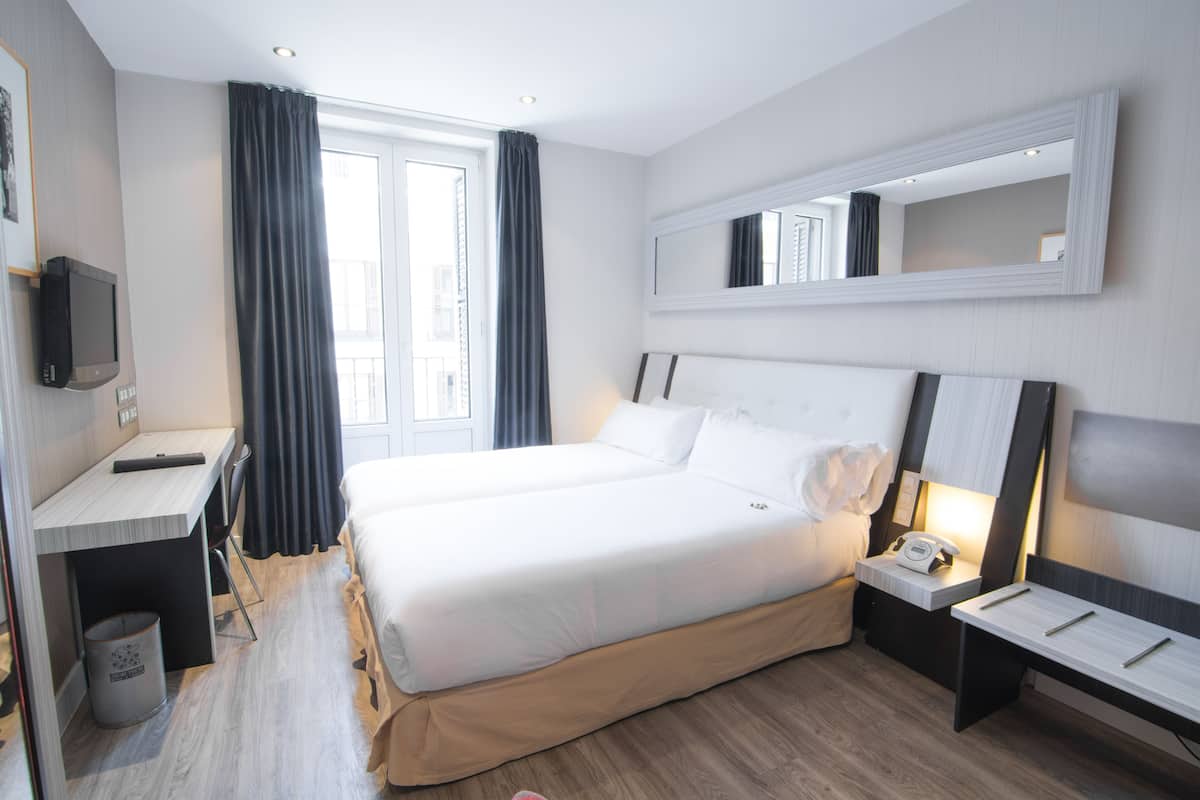Quarto de hotel com cama, mesa de cabeceira, luminária, televisão e escrivaninha com cadeira.