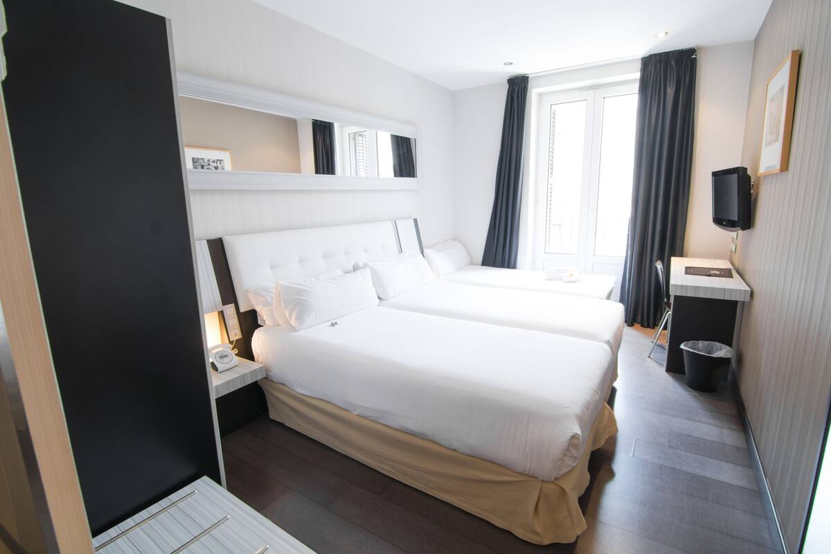 Quarto de hotel com duas camas, uma mesa de cabeceira com abajur, televisão na parede e uma janela com cortinas.