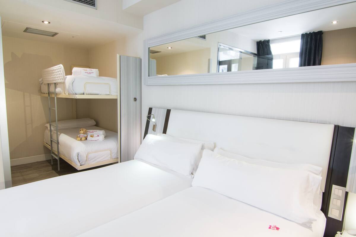 Quarto de hotel com uma cama beliche, uma cama de solteiro, um espelho grande e uma janela com cortinas.