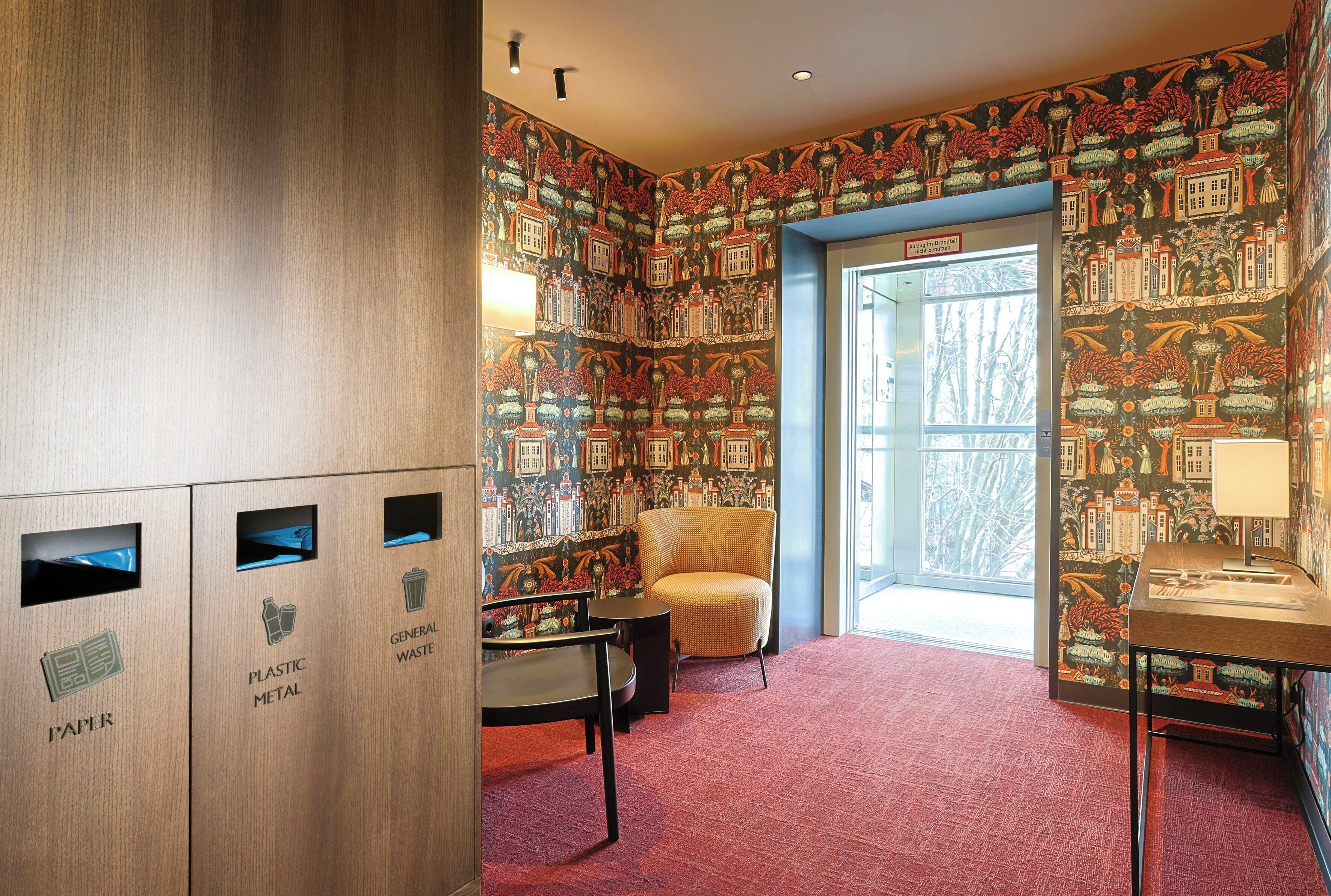 Quarto com papel de parede estampado, um armário de madeira com contentores de reciclagem, uma cadeira, uma mesa e uma janela com vista.