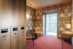 Quarto com papel de parede estampado, um armário de madeira com contentores de reciclagem, uma cadeira, uma mesa e uma janela com vista.