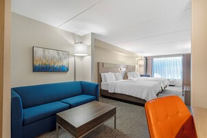Quarto de hotel com um sofá azul, uma mesa de centro de madeira, uma cama com roupa de cama branca e uma cadeira laranja vibrante.