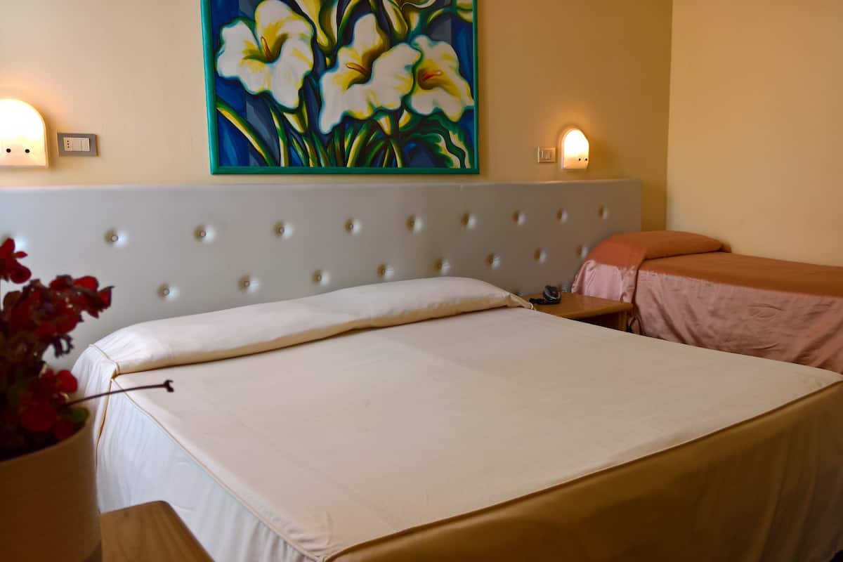 Quarto de hotel com uma cama grande, uma cama menor, uma luminária fixada na parede e um quadro de flores.