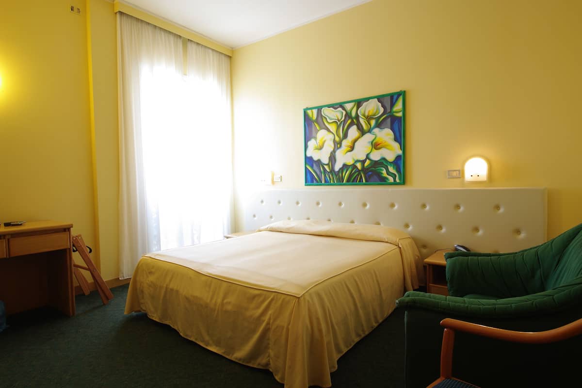 Quarto de hotel com uma cama, uma escrivaninha, uma cadeira, um quadro e uma mesinha de cabeceira.
