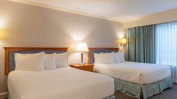 Quarto de hotel com duas camas, cada uma com cabeceira de madeira, mesa de cabeceira e luminária, e vista para uma janela com cortinas.