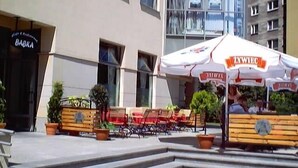 Área externa com bancos de madeira e plantas em vasos, uma tenda branca com sinalização, e um edifício com a placa "BARKA".