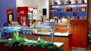 Uma área de cozinha com geladeira, máquina de refrigerantes Coca-Cola e prateleiras repletas de diversos alimentos.