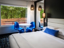 Quarto de hotel com cama, duas cadeiras azuis, uma mesinha e vista para o verde lá fora.