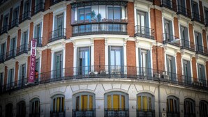 Um edifício de vários andares com estilo arquitetônico clássico, com varandas e grades pretas, e uma faixa vermelha destacada com o nome 