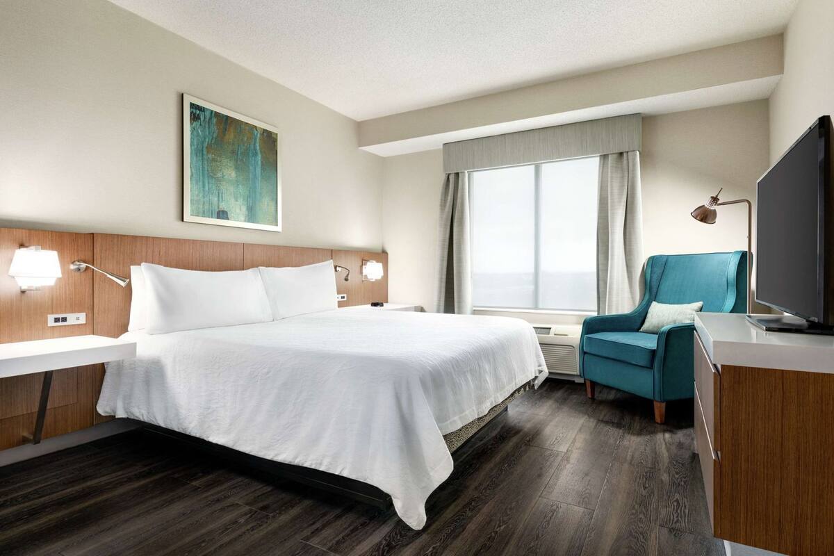 Quarto de hotel com cama, escrivaninha, televisão de tela plana, cadeira e janela com cortinas.