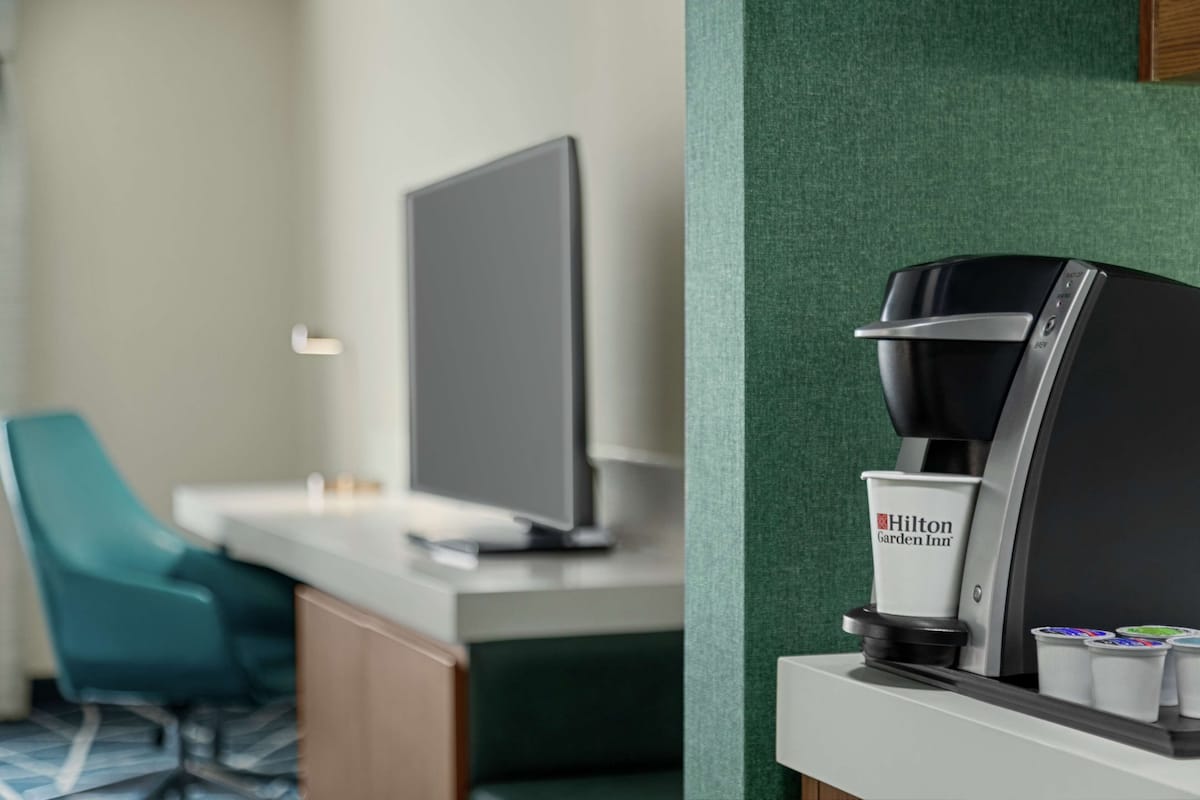 Quarto de hotel com escrivaninha, monitor de computador, máquina de café e uma xícara Hilton Garden Inn.