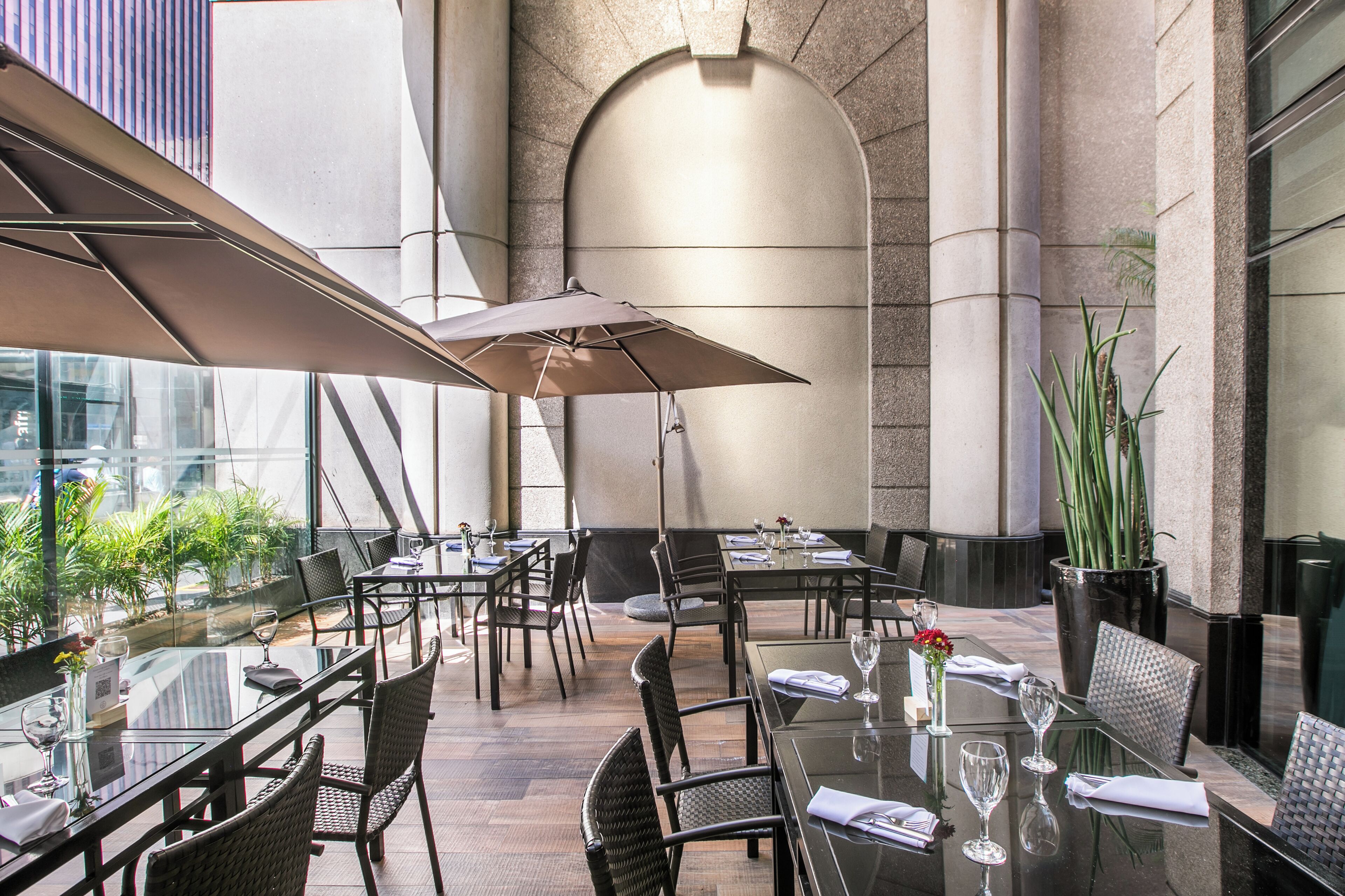 Um restaurante com mesas e cadeiras de vidro, um guarda-sol grande e um vaso com planta.