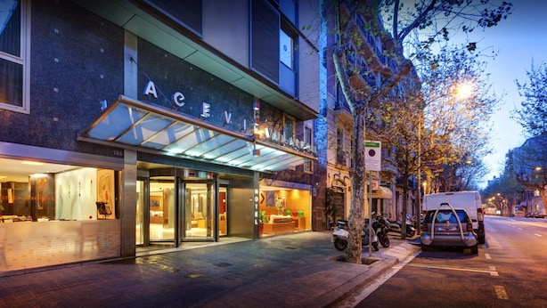 Entrada de hotel com porta de vidro, uma placa acima da porta e vista da rua com veículos estacionados e árvores.