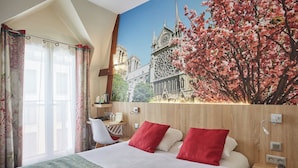 Quarto de hotel com uma cama grande, almofadas vermelhas, uma mesa com cadeira e um mural de uma catedral e flores de cerejeira.