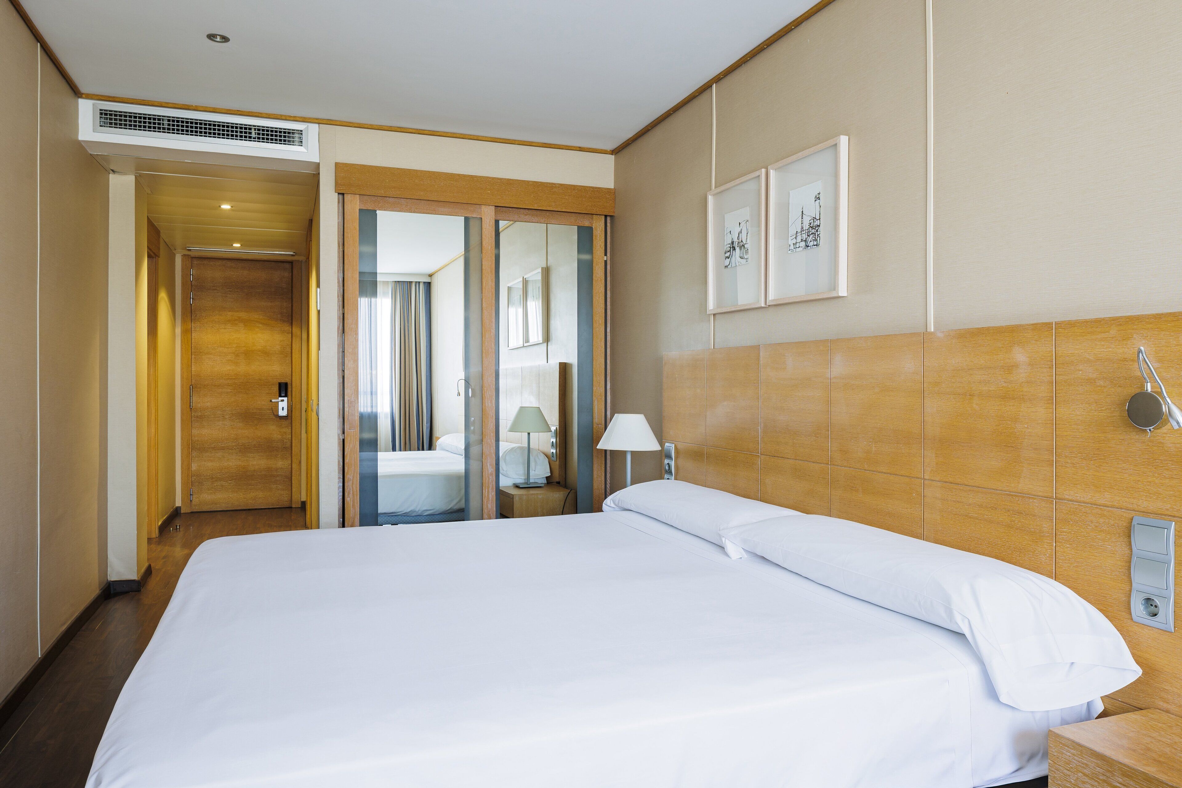 Quarto de hotel com uma cama grande, cabeceira de madeira, mesa de cabeceira com abajur e porta que dá para outro quarto.