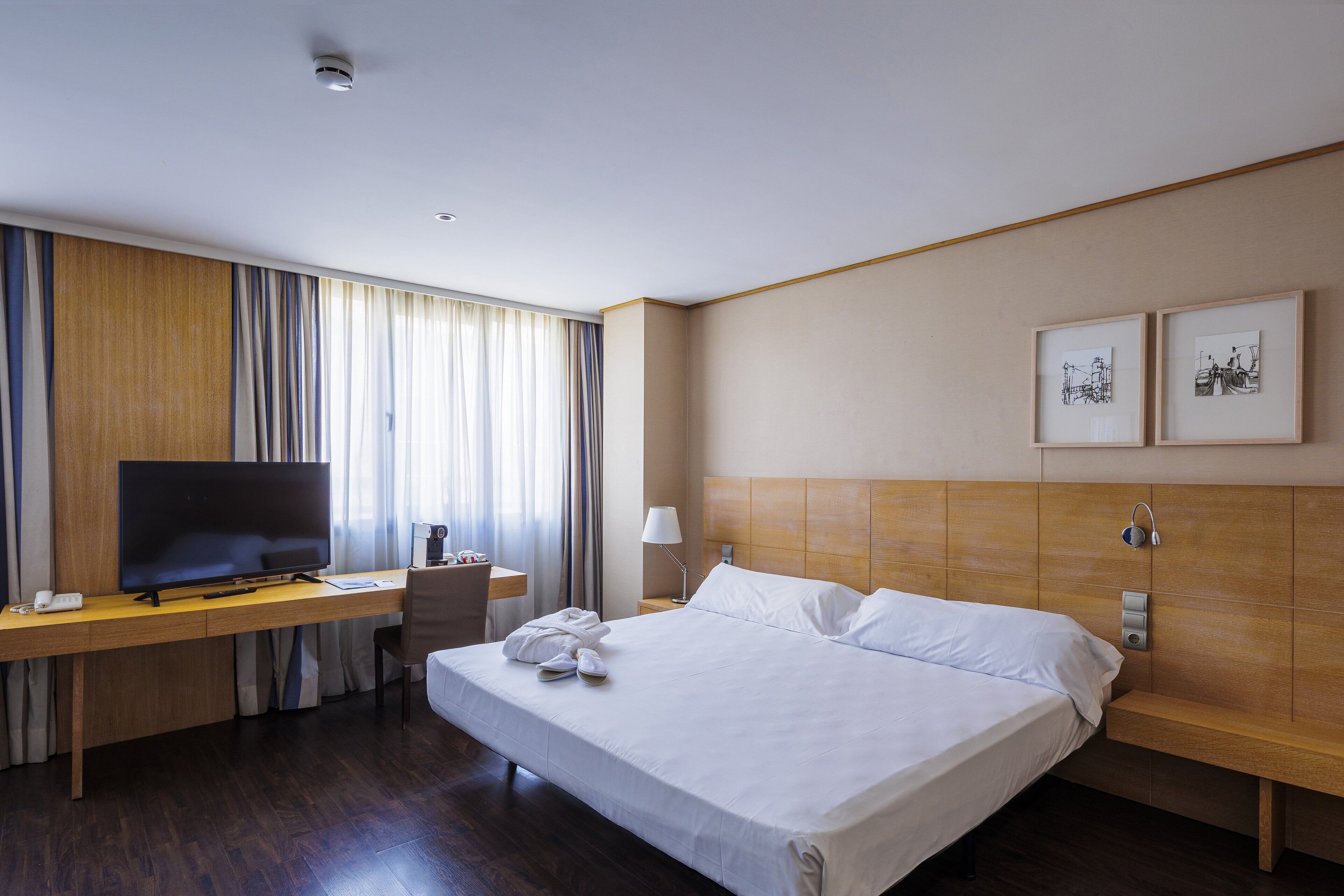 Quarto de hotel com cama, escrivaninha, televisão, cadeira e abajur.