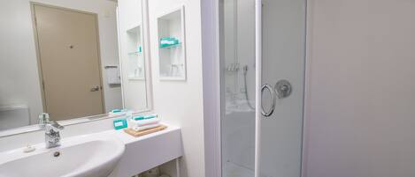 Casa de banho com lavatório branco, espelho, cabine de duche em vidro e artigos de higiene no balcão.