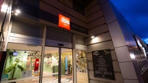 Exterior de um hotel ibis com fachada moderna, grandes janelas de vidro e uma placa vermelha chamativa.