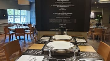 Interior de restaurante com um quadro de menu exibindo diversos pratos e preços. As mesas estão montadas com pratos, talheres e copos.